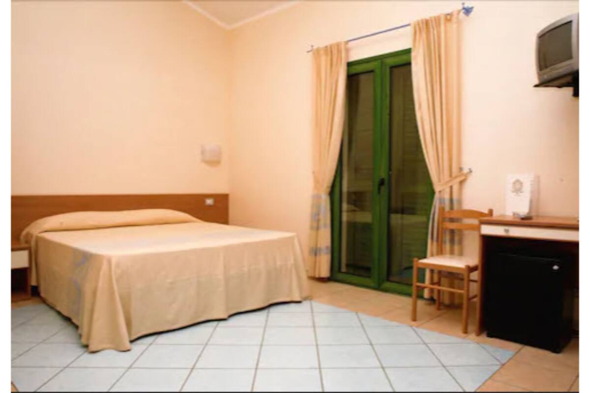 Quarto pequeno e arrumado com uma cama, uma escrivaninha, uma cadeira e uma televisão.