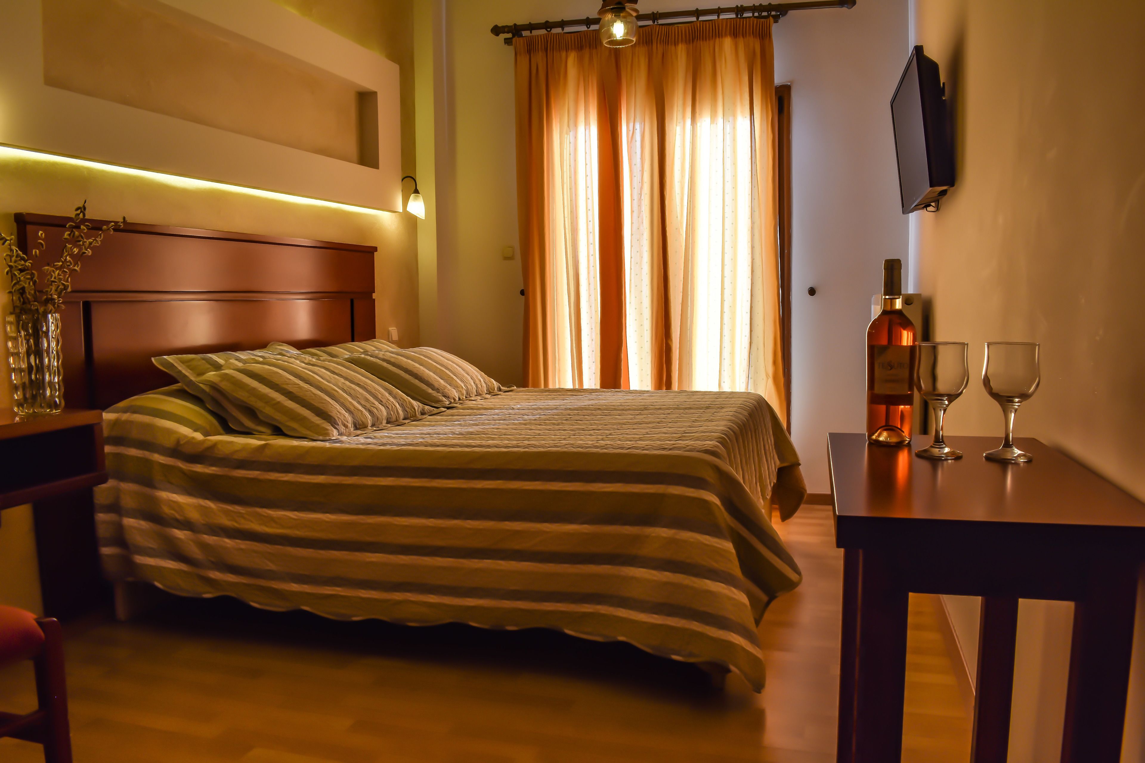Uma cama bem arrumada com colcha listrada, uma mesa de cabeceira com garrafa e copos, e uma TV fixada na parede.