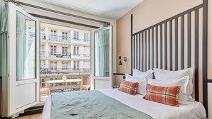 Quarto de hotel com uma cama grande, varanda com mesa e cadeiras, e vista para outros edifícios.