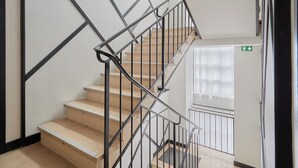 Uma escada com corrimão de metal, degraus bege e uma porta com placa de saída de emergência verde.