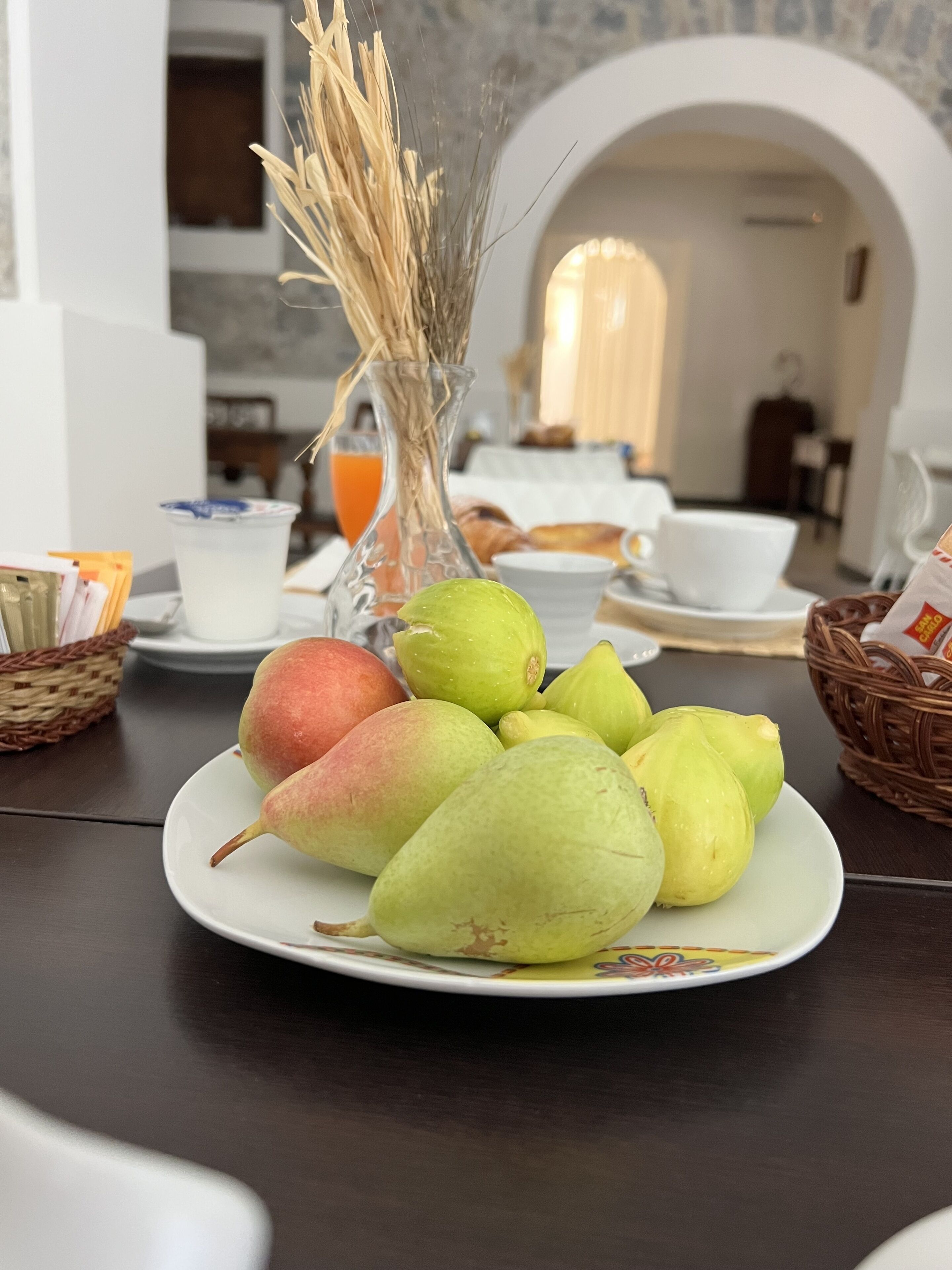 Uma mesa de jantar com um prato de frutas, um copo de suco de laranja e um vaso com plantas secas.