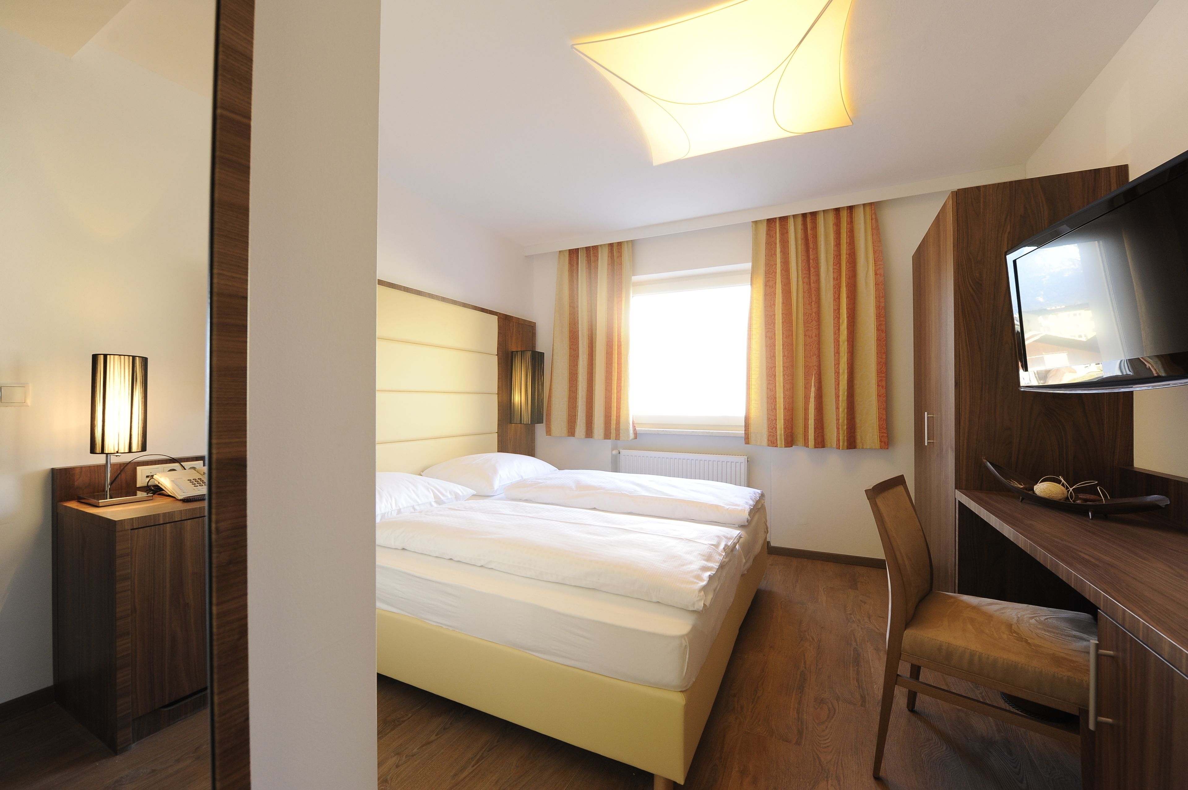Quarto de hotel com uma cama, uma escrivaninha com televisão, uma cadeira e uma mesa de cabeceira com abajur.