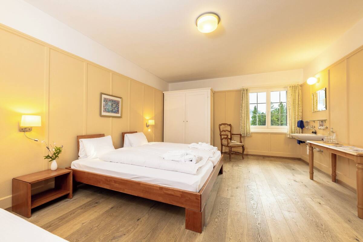 Quarto de hotel com cama de madeira, mesas de cabeceira, uma cadeira, janela com cortinas, pia e espelho.