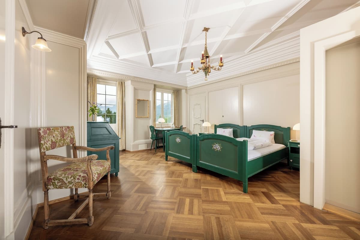 Um quarto espaçoso com piso de parquet de madeira, cama estofada em verde, uma cadeira de madeira, uma escrivaninha e uma janela com vista.