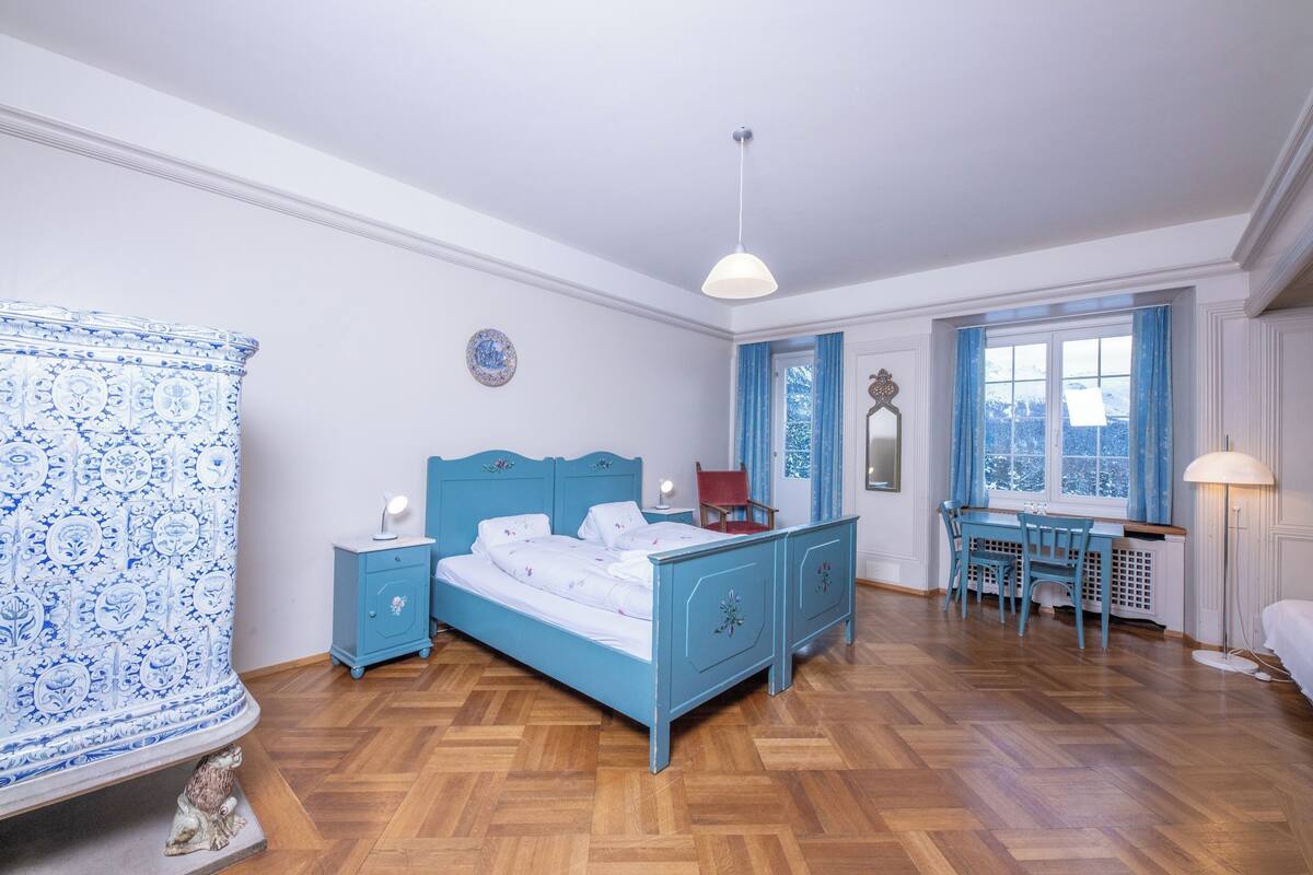 Quarto com cama azul, uma pequena escrivaninha, uma cadeira, abajur e janela com cortinas.