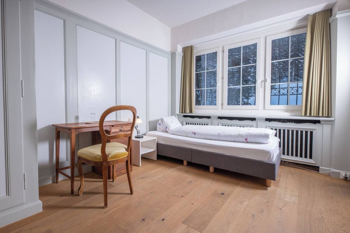 Quarto com cama, cadeira de madeira, escrivaninha e janela com cortinas.