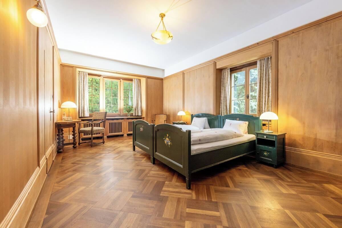 Quarto com piso de madeira, cama com cabeceira, duas mesas de cabeceira com luminárias, uma cadeira e janela com cortinas.