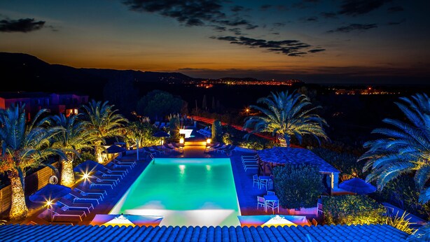 Vista noturna de um resort com piscina, espreguiçadeiras e palmeiras.