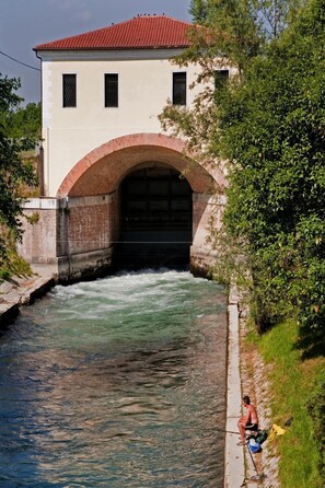 Um rio correndo sob uma ponte, com uma pessoa pescando na margem.