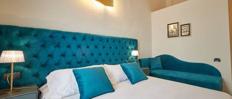 Quarto de hotel com uma cama grande, cabeceira estofada, duas mesas de cabeceira com luminárias, um sofá azul-esverdeado e quadros nas paredes.