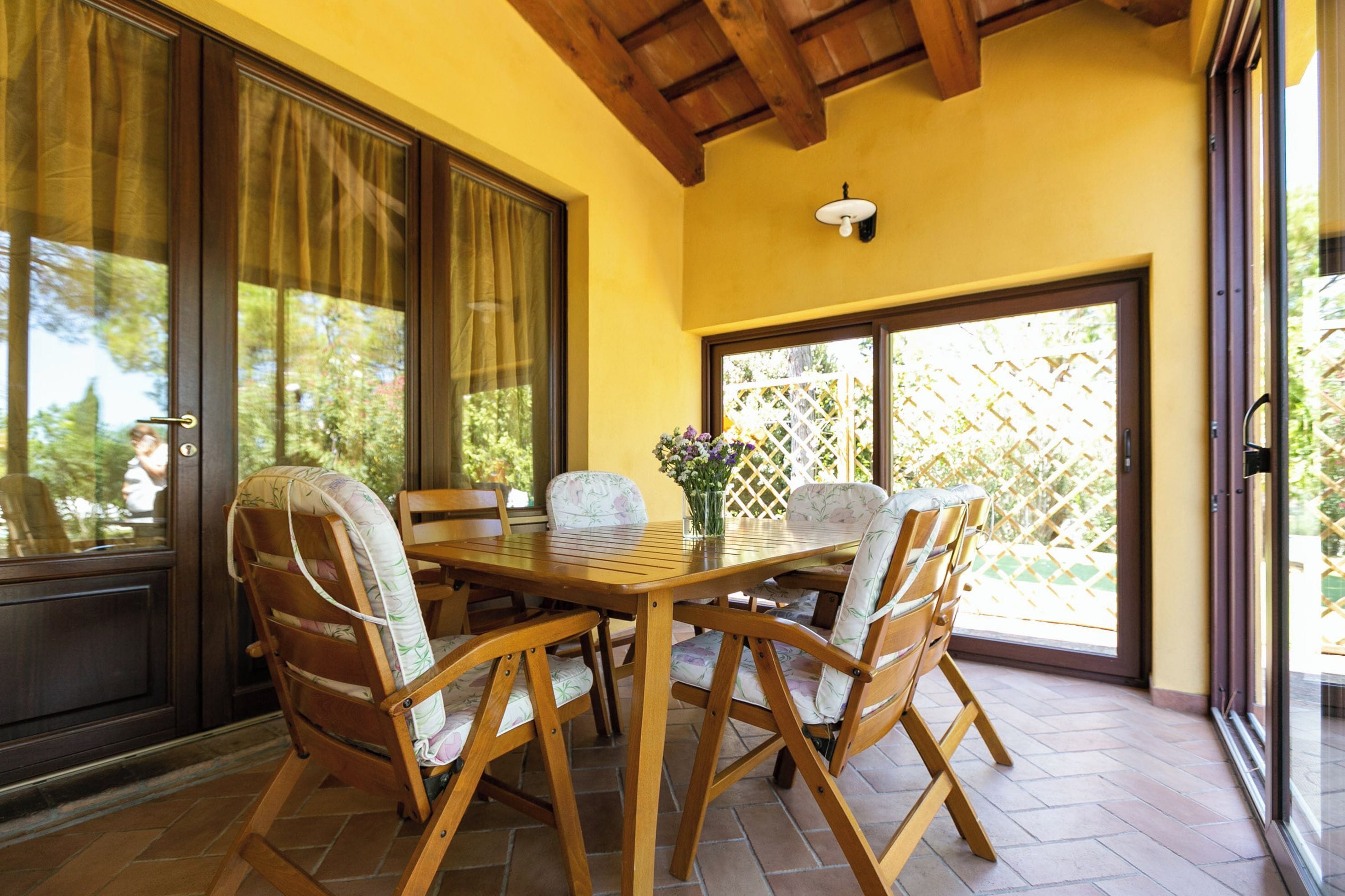 Uma mesa de jantar de madeira com quatro cadeiras, um vaso com flores e vista para o verde através de grandes janelas.