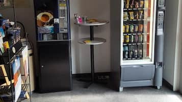 Quarto com máquina de vendas, uma pequena mesa e uma estante de revistas.