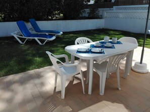 Uma área de pátio com uma mesa branca posta para refeições, rodeada por cadeiras de plástico branco.