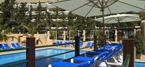 Piscina com espreguiçadeiras azuis e guarda-sóis, cercada por palmeiras e edifícios.