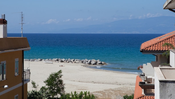Vista costeira com praia arenosa, molhe rochoso e edifícios com varandas de frente para o mar.