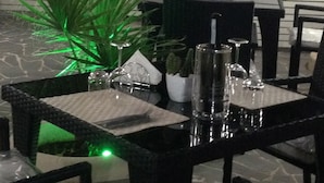 Uma área de refeições ao ar livre com mesas quadradas, cadeiras e plantas em vasos.