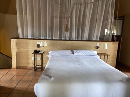 Quarto de hotel com uma cama grande, duas mesas de cabeceira com abajures, uma pequena escrivaninha com telefone e um espelho na parede.