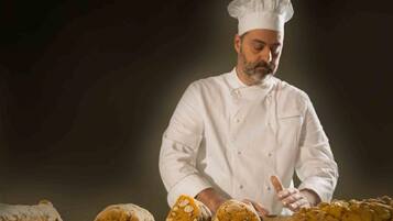 Um chef com uniforme branco e chapéu de chef, posicionado atrás de uma mesa com diversos alimentos, incluindo um pão e um doce com nozes visíveis.