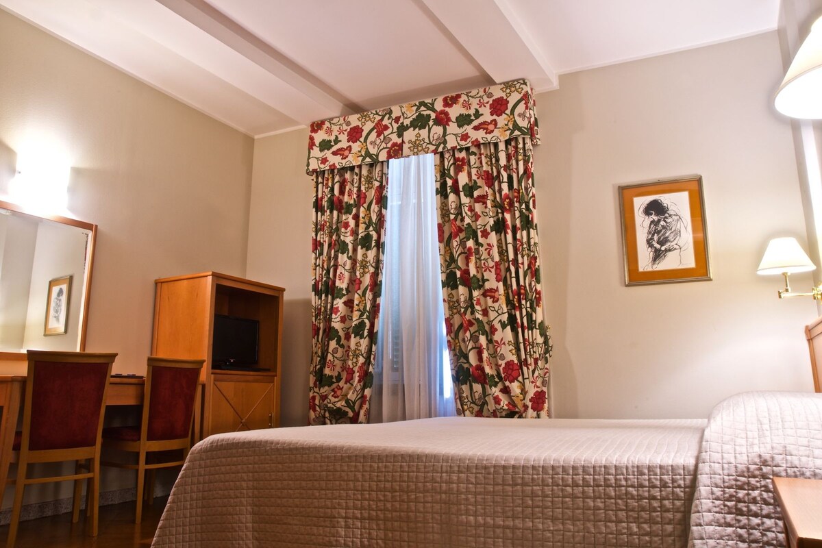 Quarto de hotel com uma cama, uma escrivaninha com cadeira, uma televisão e uma janela com cortinas florais.