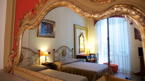Quarto de hotel com duas camas, um espelho grande e trabalhado, uma escrivaninha com luminária e vista para o exterior através de uma janela com cortinas.