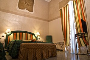 Quarto com cama, duas luminárias fixadas na parede, cabeceira estofada verde, colcha estampada e janela com cortinas listradas.
