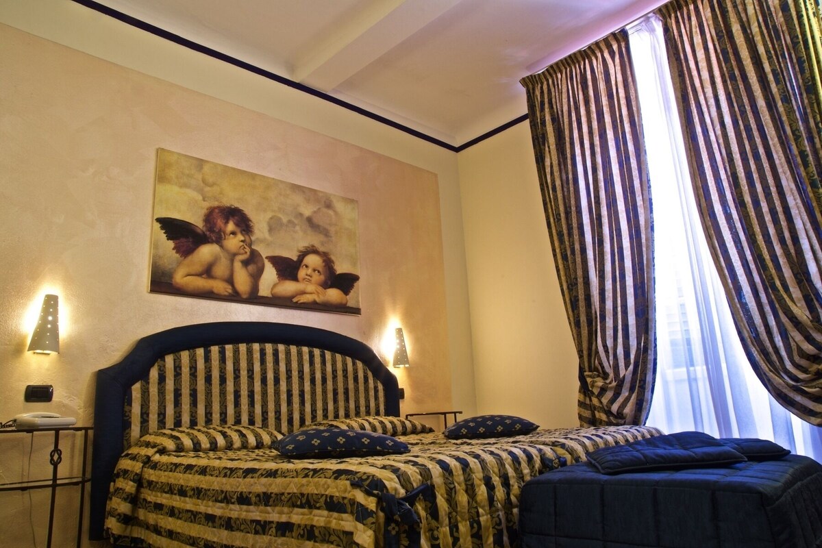 Quarto de hotel com cama, mesa de cabeceira, abajur e cortinas.