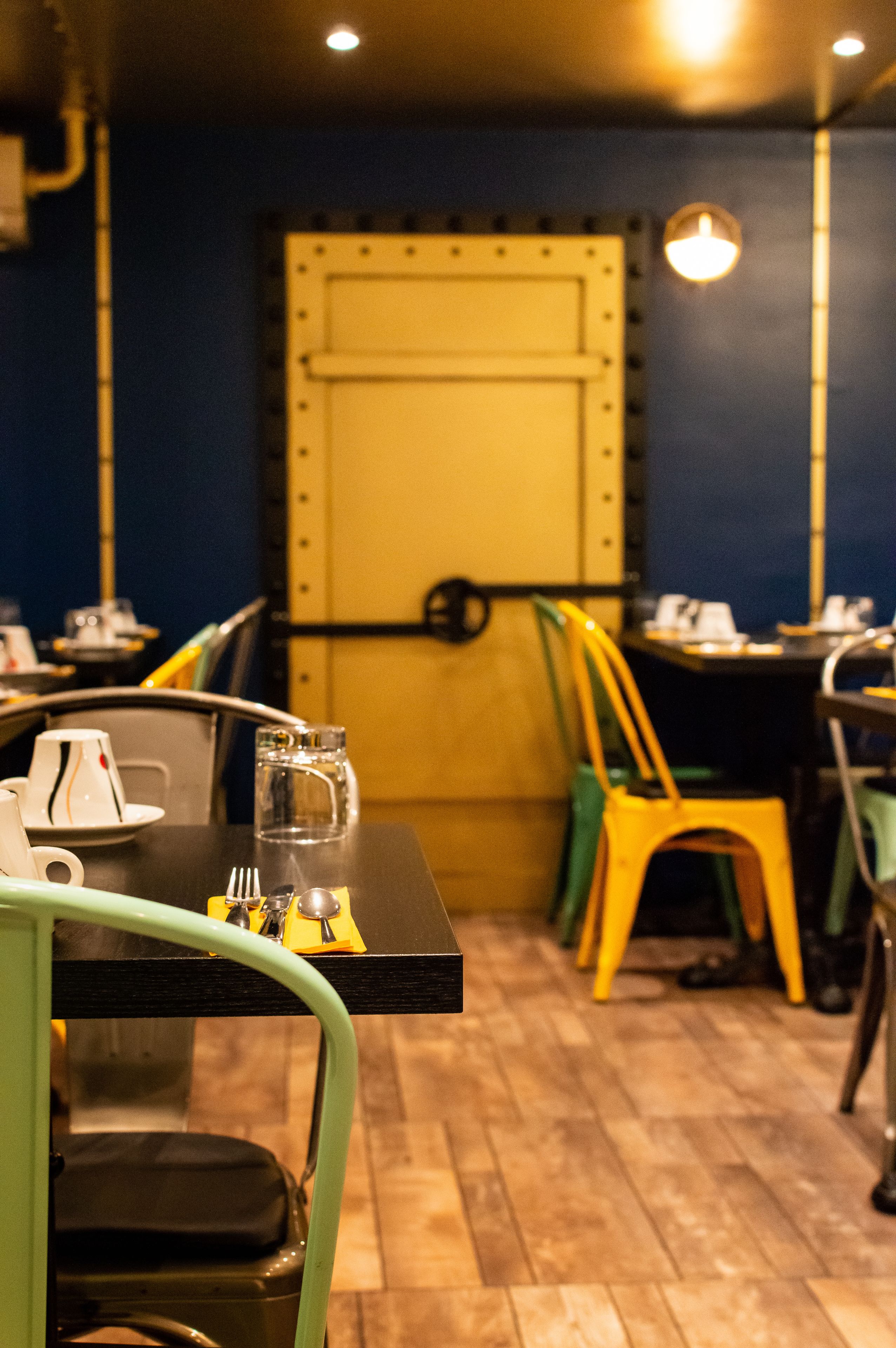 Interior de restaurante com chão de madeira, cadeiras amarelas e verdes, uma mesa posta com xícara, copo e talheres.