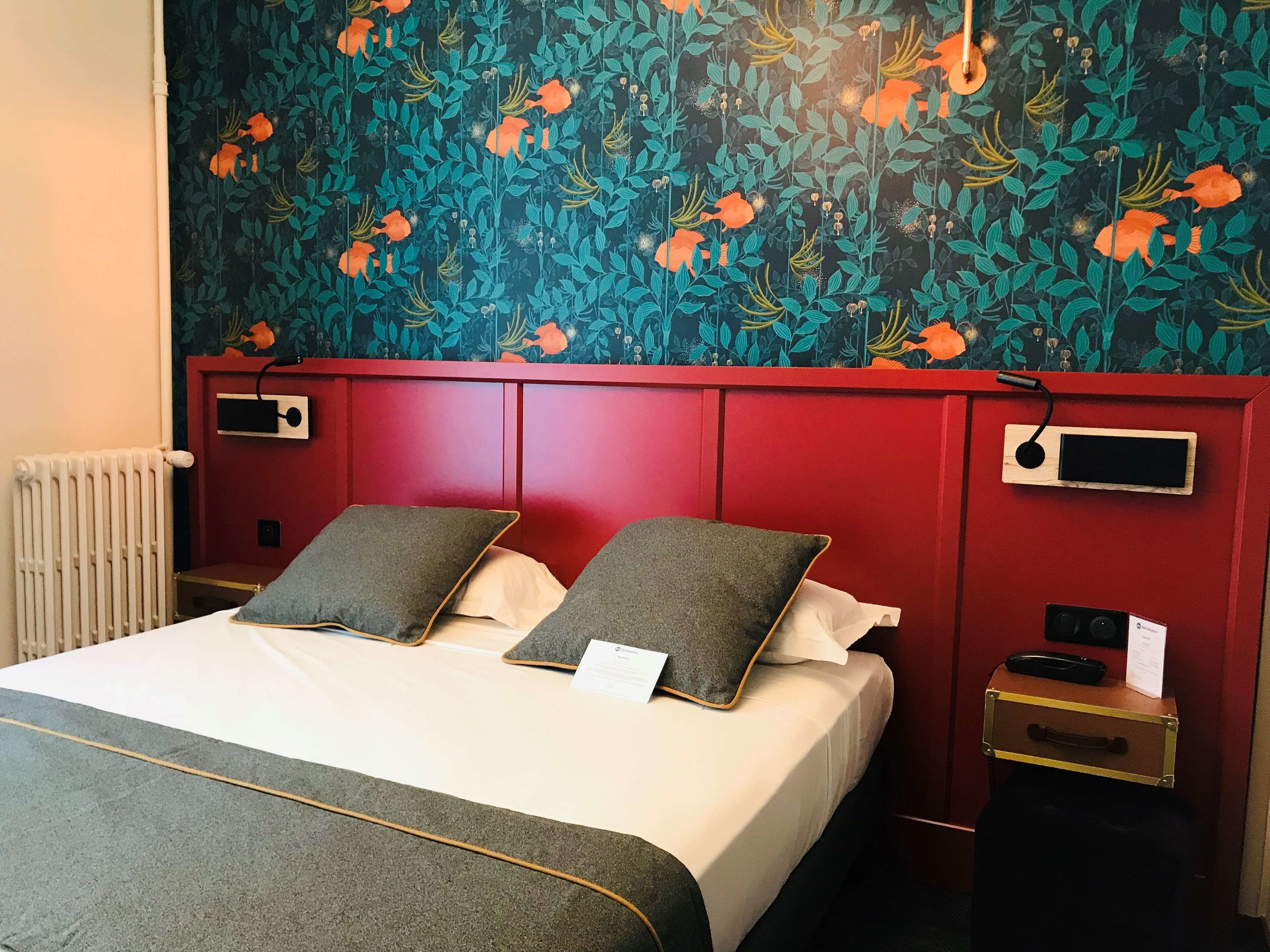 Quarto de hotel com cama, duas almofadas, mesa de cabeceira, cabeceira vermelha e papel de parede floral.