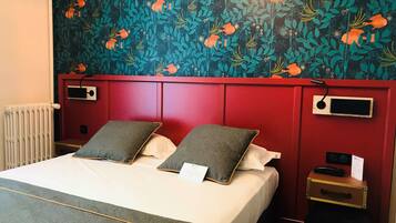 Quarto de hotel com cama, duas almofadas, mesa de cabeceira, cabeceira vermelha e papel de parede floral.
