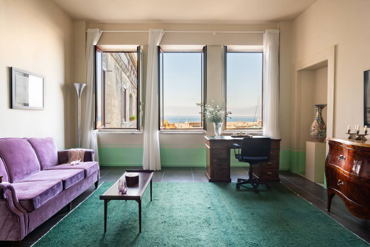 Quarto com sofá roxo, uma escrivaninha e vista para o mar através de janelas amplas.