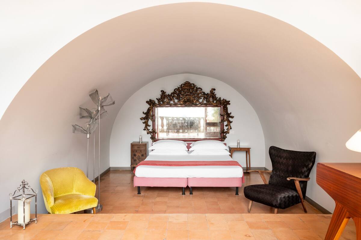 Um quarto com um teto abobadado grande, uma cama com cabeceira decorativa, uma poltrona amarela, uma poltrona preta e uma mesa de cabeceira com uma luminária.