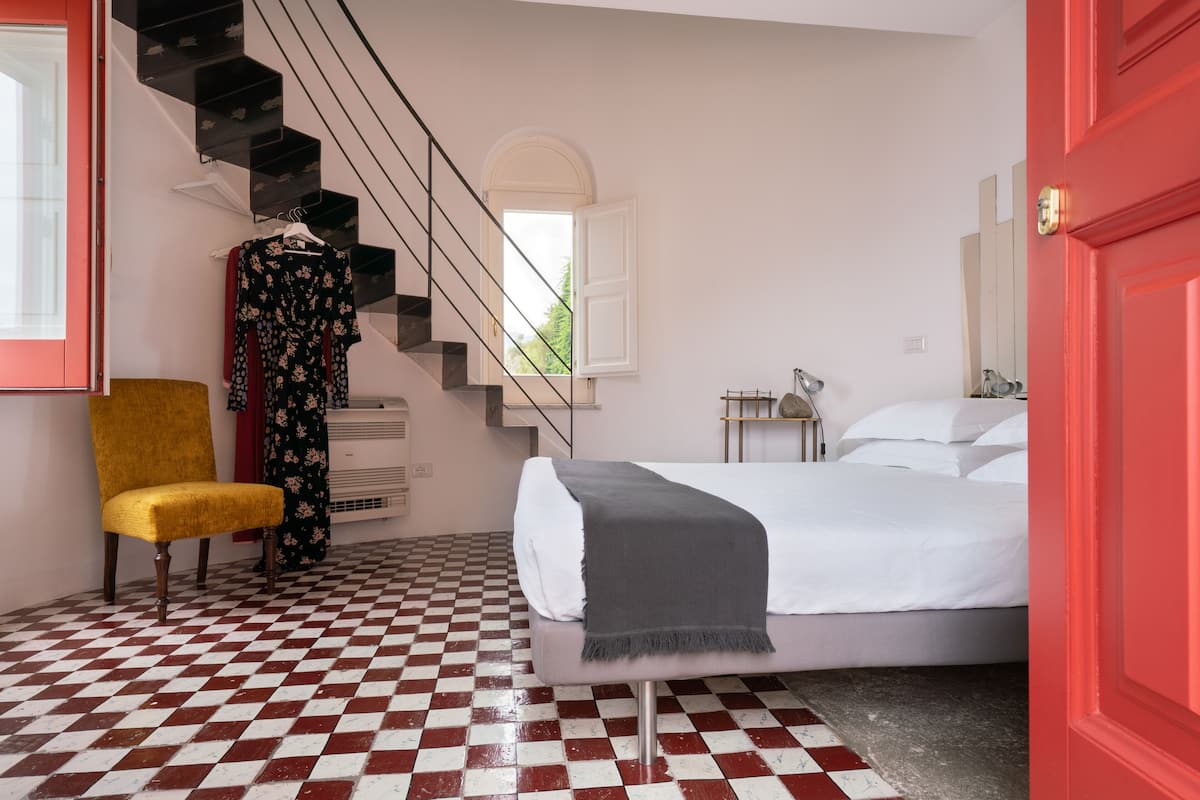 Quarto com piso xadrez, cama com roupa de cama branca, uma cadeira amarela, um vestido preto em um cabide e uma escada.