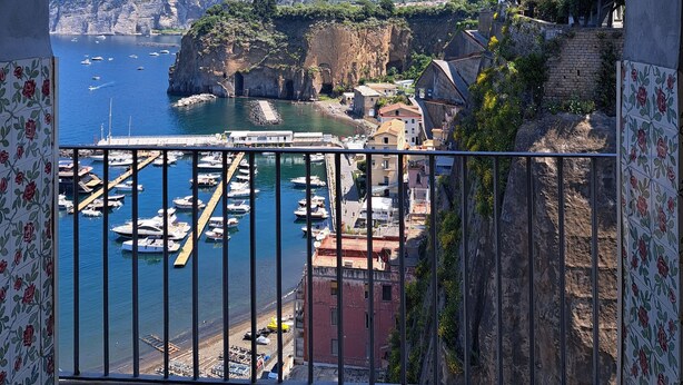 Vista costeira através de uma janela em arco com chão de azulejos, com vista para uma marina com barcos, falésias rochosas e um morro com edifícios.
