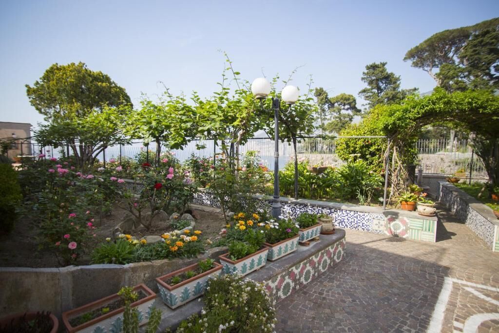 Um jardim no terraço com plantas em vasos, um caminho de azulejos e vista para o mar.