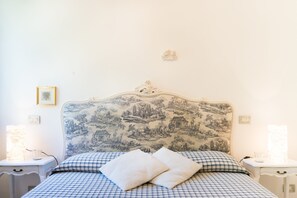 Quarto com colcha xadrez, cabeceira decorativa com cena campestre e abajures laterais.