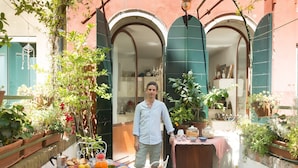 Um homem está num pátio com uma mesa posta para o pequeno-almoço, rodeado por plantas em vasos e um edifício rústico com portadas verdes.