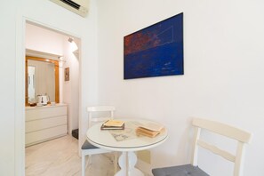 Um quarto pequeno e bem iluminado, com uma mesa redonda, uma cadeira branca e um quadro na parede.