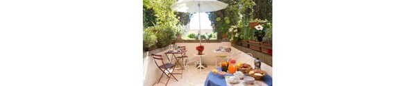Pátio externo com mesa posta para refeição, cercado por vasos de plantas e um guarda-sol branco.