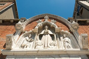 Escultura arquitetônica em pedra com figuras e uma figura central, em frente a uma parede de tijolos.