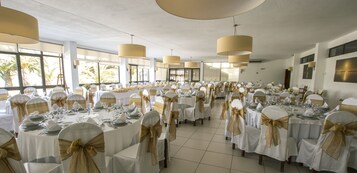 Salão de festas com mesas redondas dispostas para um evento formal, com toalhas brancas e fitas douradas nas cadeiras.