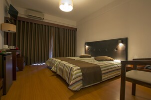 Quarto de hotel com cama, mesa de cabeceira, luminária e televisão.