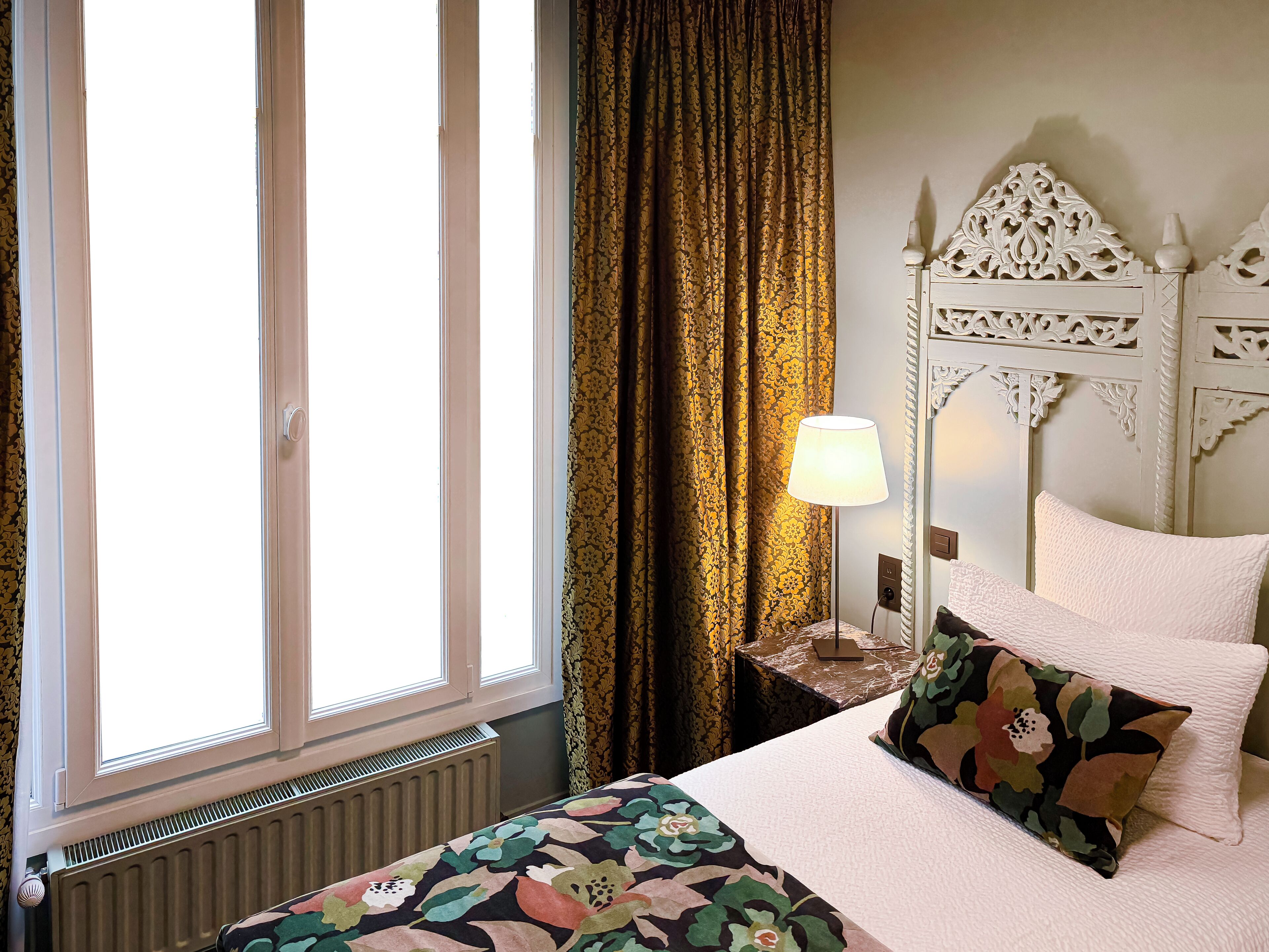 Quarto de hotel com cama, mesa de cabeceira, abajur e janela com cortinas.