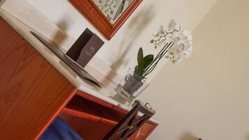 Quarto de hotel com escrivaninha de madeira, espelho, vaso com flores, telefone e mesa de cabeceira pequena.