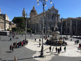 Uma praça pública espaçosa com um edifício histórico imponente, arquitetura clássica, uma fonte central e uma estátua.