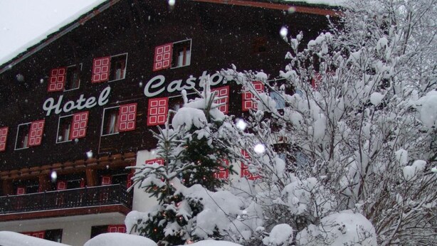 Placa do Hotel Castor coberta de neve, com base de pedra, rodeada por árvores carregadas de neve e um edifício com janelas emolduradas a vermelho ao fundo.