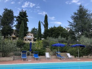 Área de piscina com espreguiçadeiras e guarda-sóis, cercada por árvores altas e um edifício de pedra ao fundo.