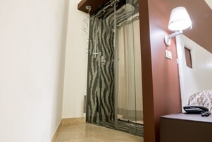 Quarto de hotel com cabine de chuveiro de vidro, mesa de cabeceira com telefone e luminária fixa na parede.