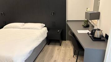 Quarto de hotel moderno com uma cama, uma escrivaninha, uma cadeira, uma mesinha, uma chaleira e uma planta em vaso.
