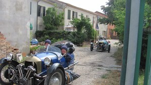 Carro antigo estacionado em um caminho de cascalho, com um edifício ao fundo.