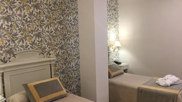 Quarto de hotel com duas camas, papel de parede floral, uma mesa de cabeceira com abajur e um espelho.